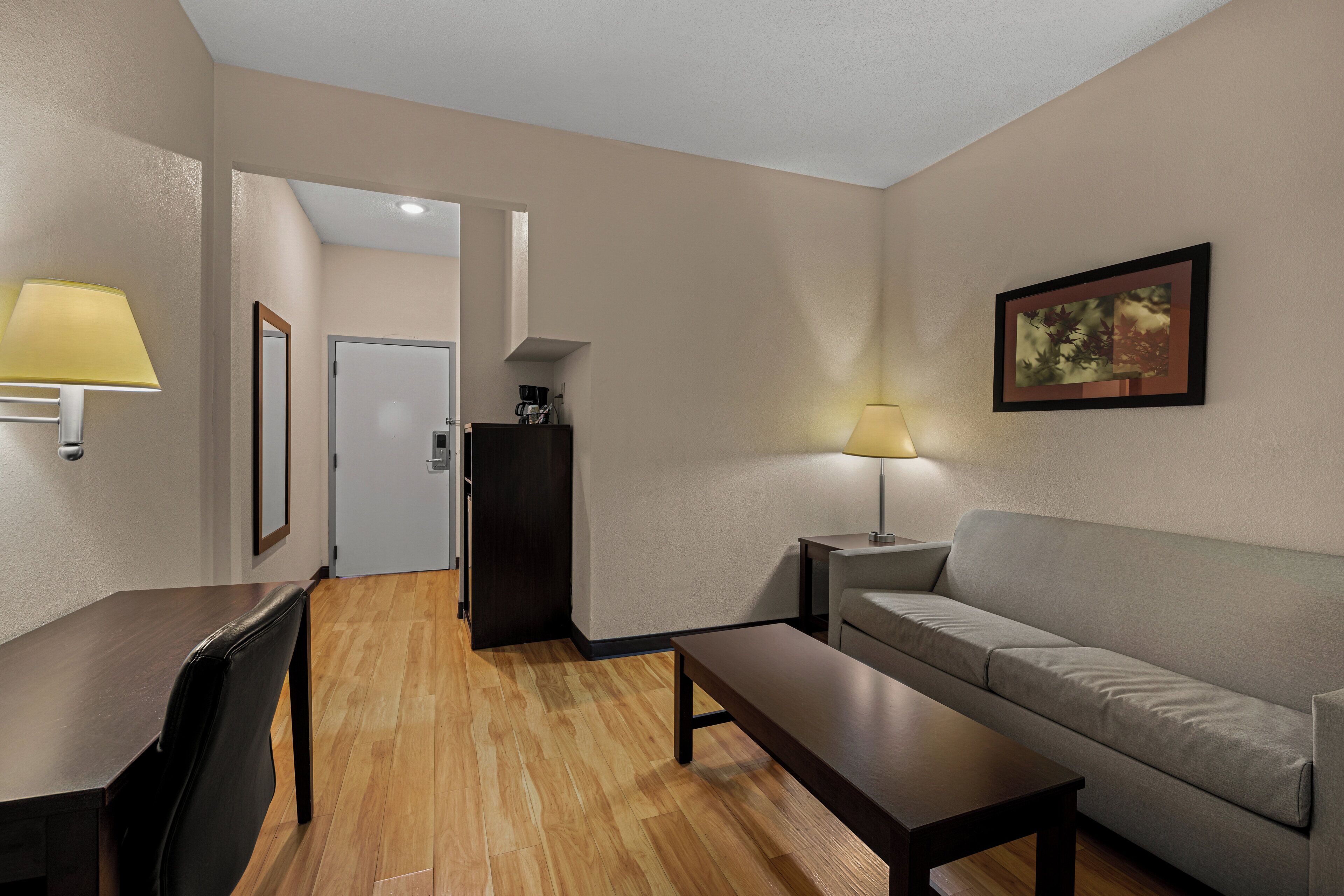 Quarto de hotel com sofá, mesa, luminária, pintura e porta para uma cozinha compacta.