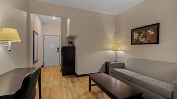 Quarto de hotel com sofá, mesa, luminária, pintura e porta para uma cozinha compacta.
