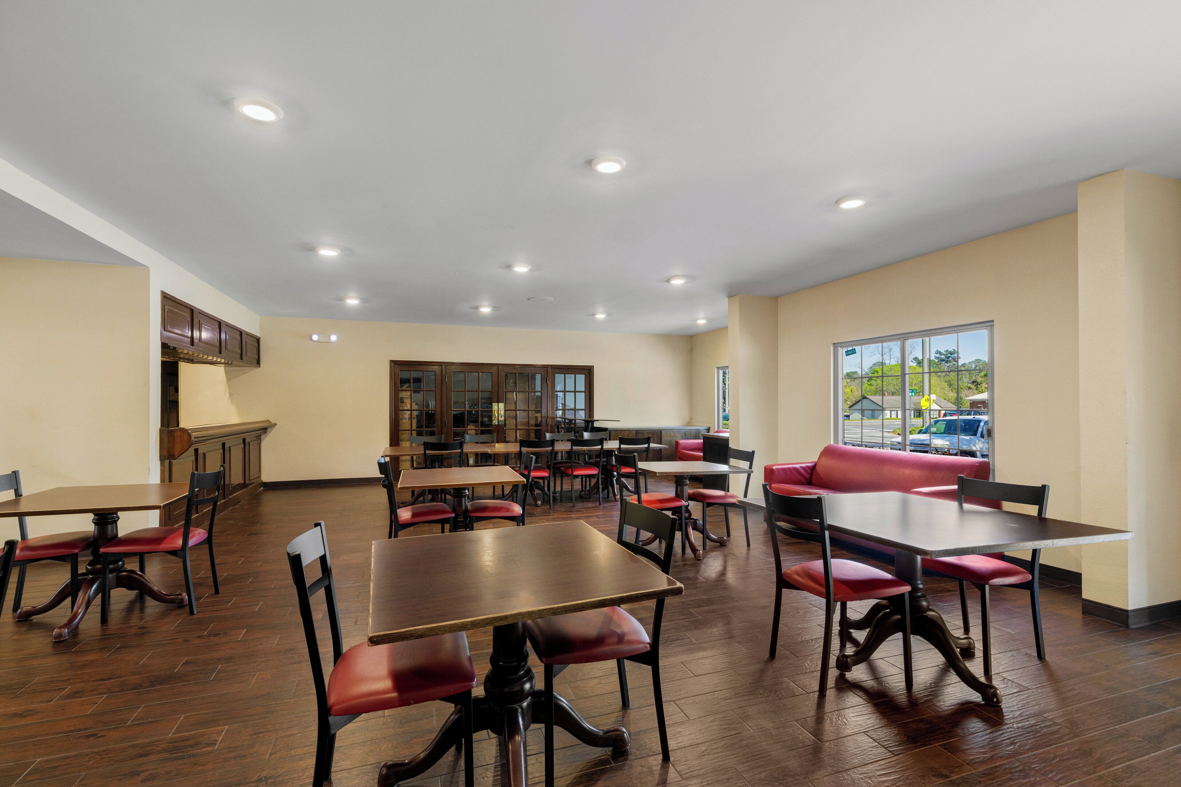 Uma ampla área de refeições interna com várias mesas e cadeiras, uma área de cozinha ao fundo e uma grande janela com vista para o exterior.