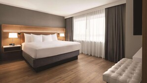 Quarto de hotel moderno com uma cama grande, abajures, uma cadeira e uma janela ampla com cortinas.