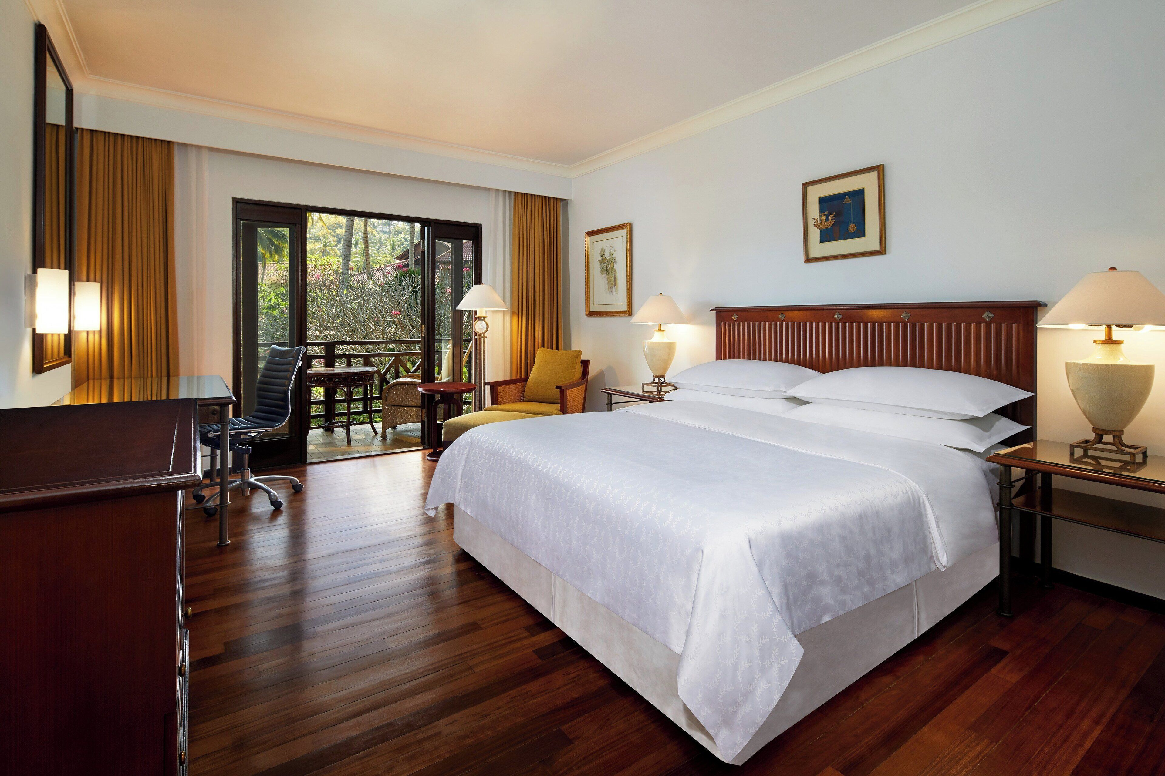 Quarto de hotel com cama grande, escrivaninha, cadeira e varanda com vista para vegetação.