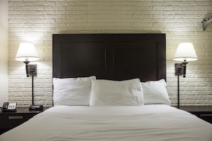 Quarto de hotel com parede de tijolos, cabeceira escura, duas luminárias de cabeceira, telefone e cama bem arrumada com lençóis brancos.