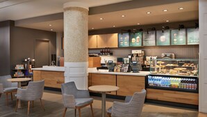 Um café moderno com uma mesa redonda central, cadeiras estofadas cinzas, um balcão de café e uma vitrine com doces.