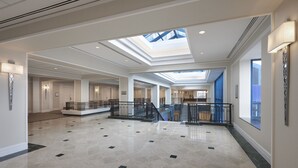 Um hall de hotel espaçoso, com teto alto, claraboias e uma escadaria central.