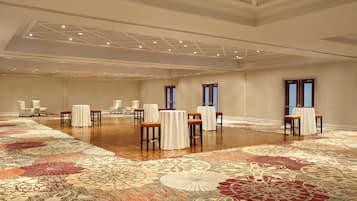 Uma sala de conferências ampla com carpete estampado, várias mesas com cadeiras e grandes janelas que permitem entrada de luz natural.