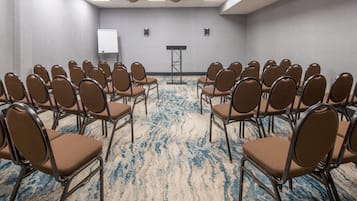 Uma sala de conferências com fileiras de cadeiras voltadas para um púlpito, uma lousa branca e dois alto-falantes nas paredes.