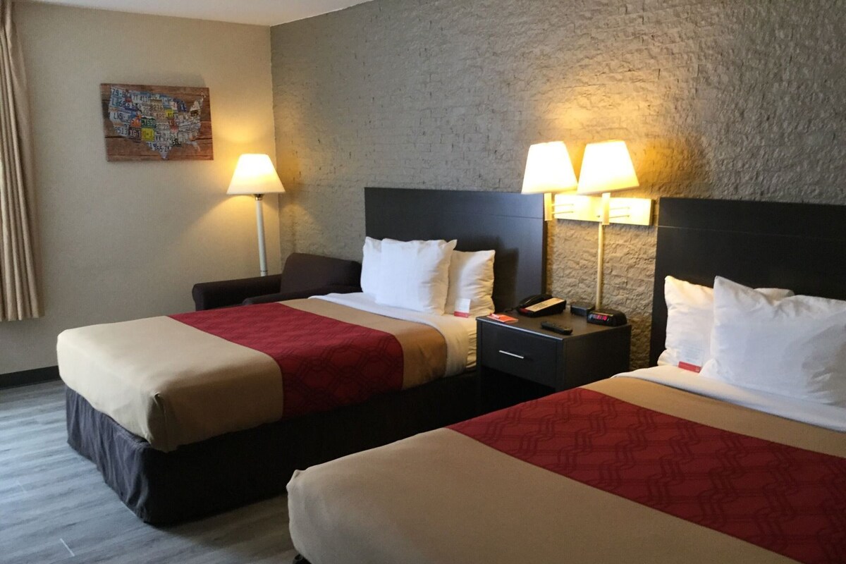 Quarto de hotel com duas camas, cada uma com colcha vermelha e bege, uma mesa de cabeceira com abajur, e uma luminária na parede.