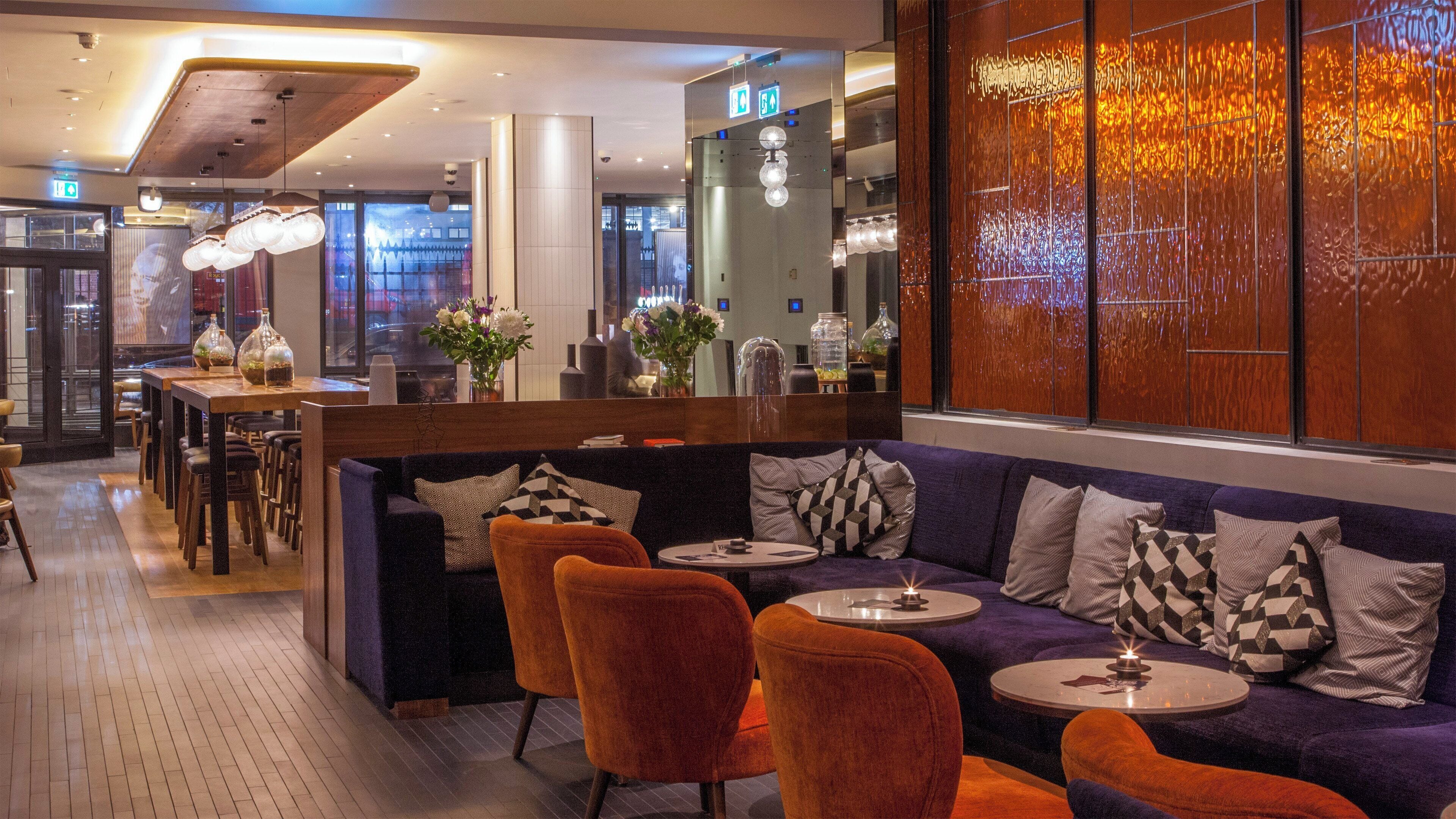 Espaço moderno com área de estar com sofás e cadeiras, bar com balcão e parede decorativa com superfície refletora.