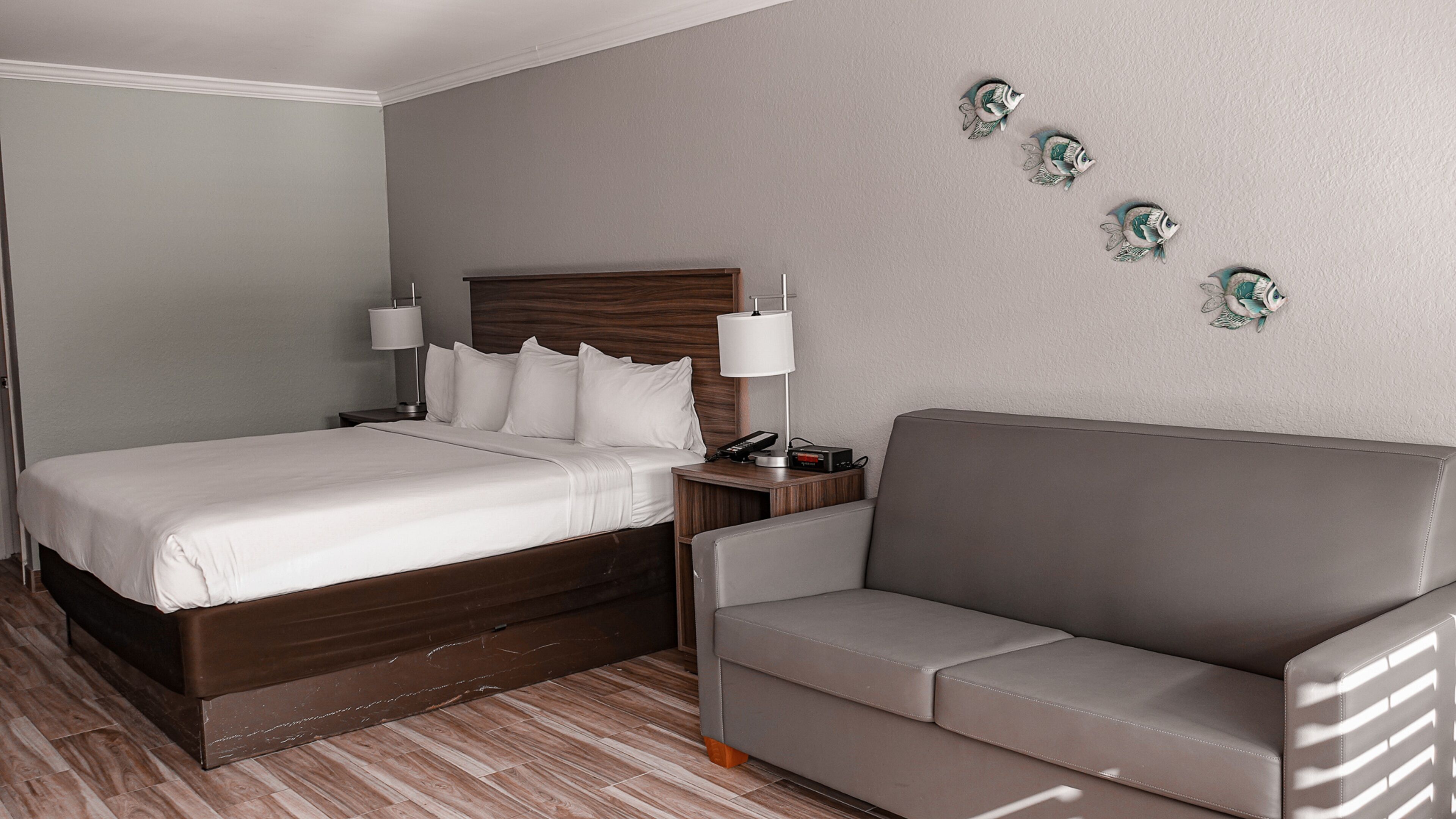 Quarto de hotel com cama, mesa de cabeceira com abajur, controle remoto e sofá cinza.