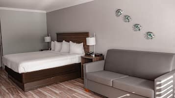 Quarto de hotel com cama, mesa de cabeceira com abajur, controle remoto e sofá cinza.