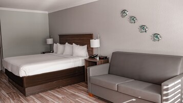 Quarto de hotel com cama, mesa de cabeceira com abajur, controle remoto e sofá cinza.