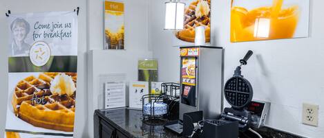 Espaço para o café da manhã com máquinas de waffle, cafeteira e um aviso convidando os hóspedes a participar.