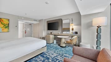 Quarto de hotel com cama, escrivaninha, poltrona, sofá, televisão, espelho, luminária e tapete estampado.