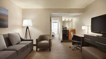 Quarto de hotel com sofá, poltrona, escrivaninha, televisão e área de kitchenette.