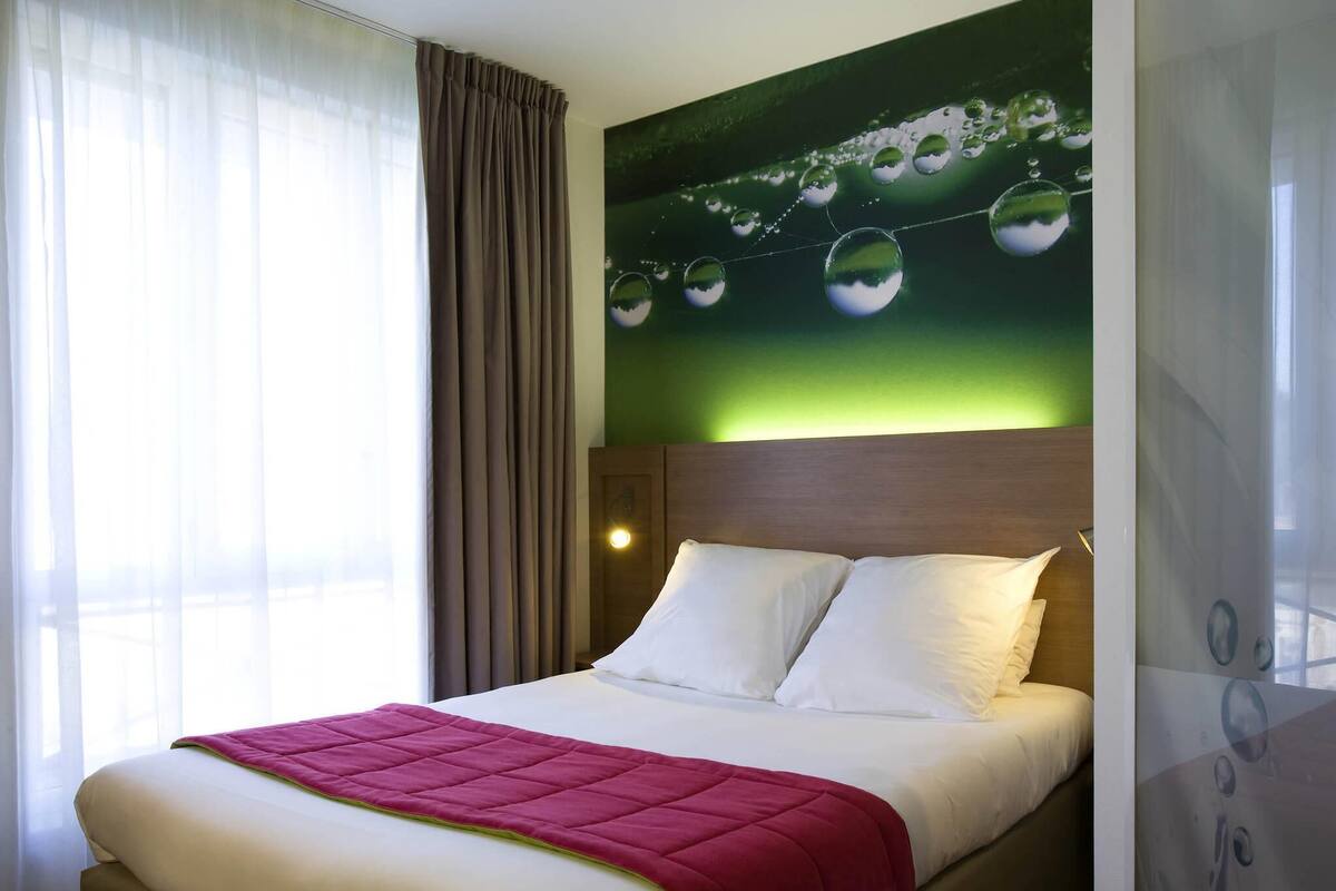 Quarto de hotel com uma cama, uma janela com cortinas, uma parede com um mural verde e uma luz noturna.