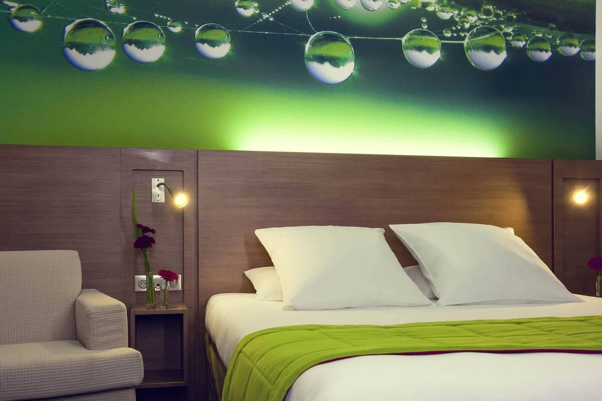 Quarto de hotel com uma cama, uma cadeira, uma mesa de cabeceira e um espelho fixado na parede.