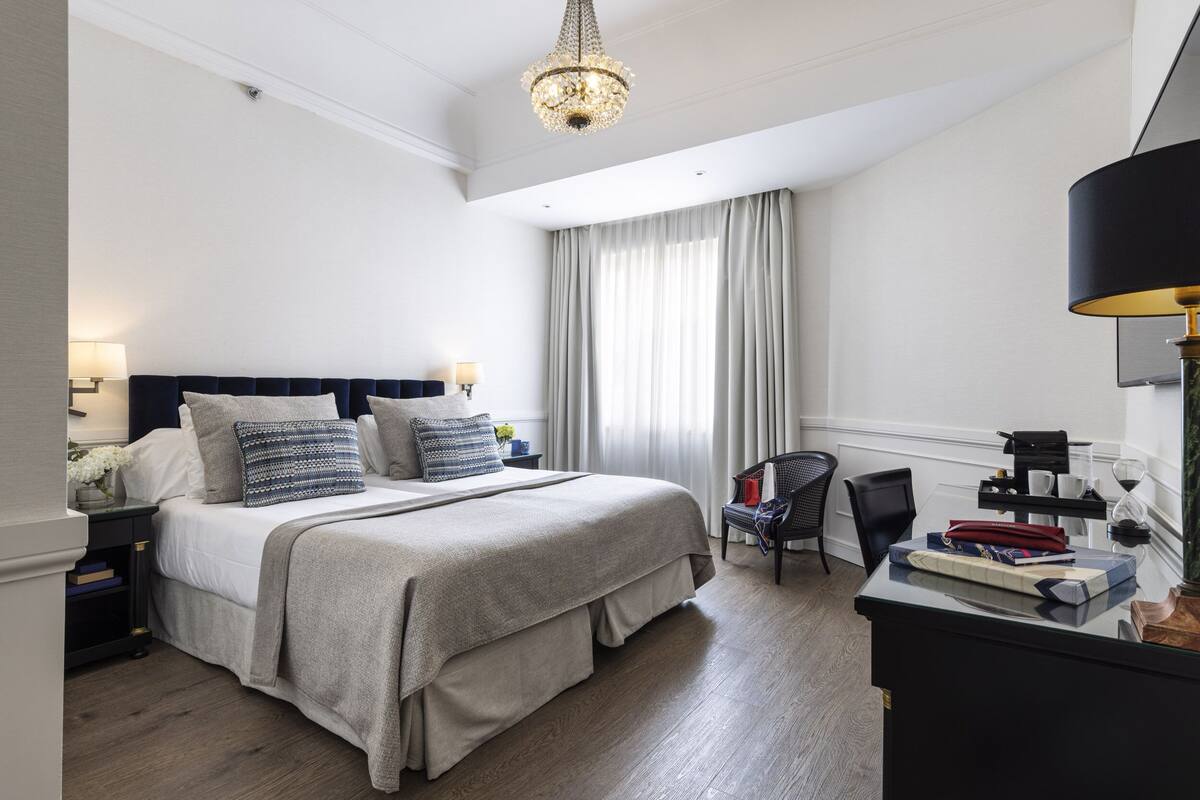 Quarto de hotel moderno com uma cama grande, uma escrivaninha com abajur e uma cadeira.