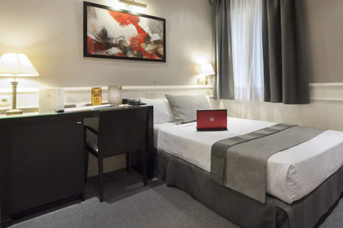 Quarto de hotel com cama, escrivaninha, laptop e mala vermelha.