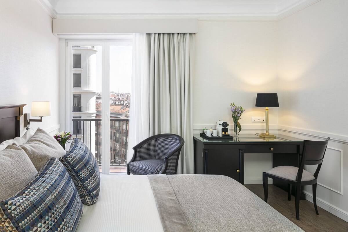 Quarto de hotel com cama, escrivaninha, cadeira e vista da cidade pela janela.