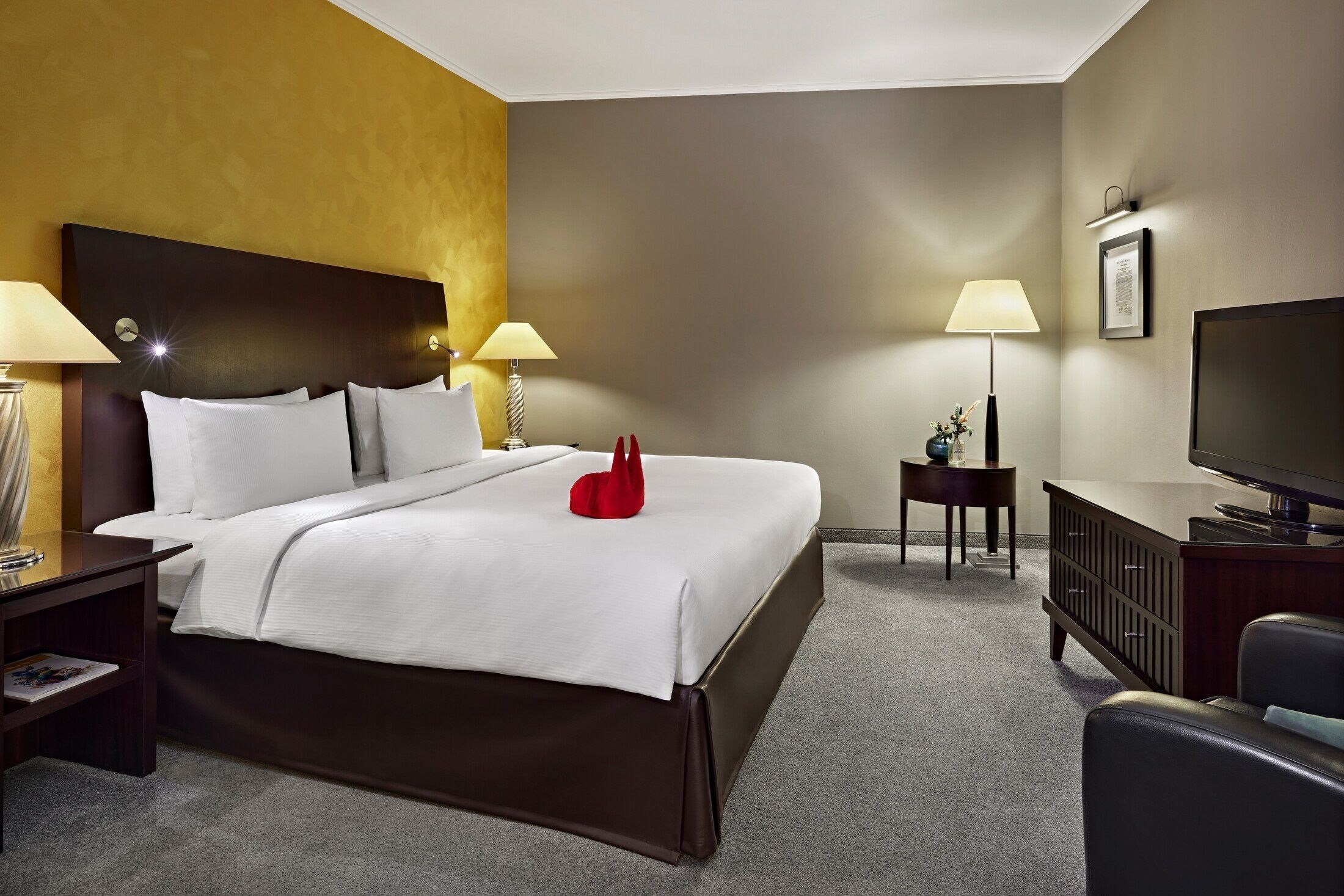 Quarto de hotel com uma cama grande, duas mesas de cabeceira com abajures, uma televisão e uma mesinha lateral com um vaso.