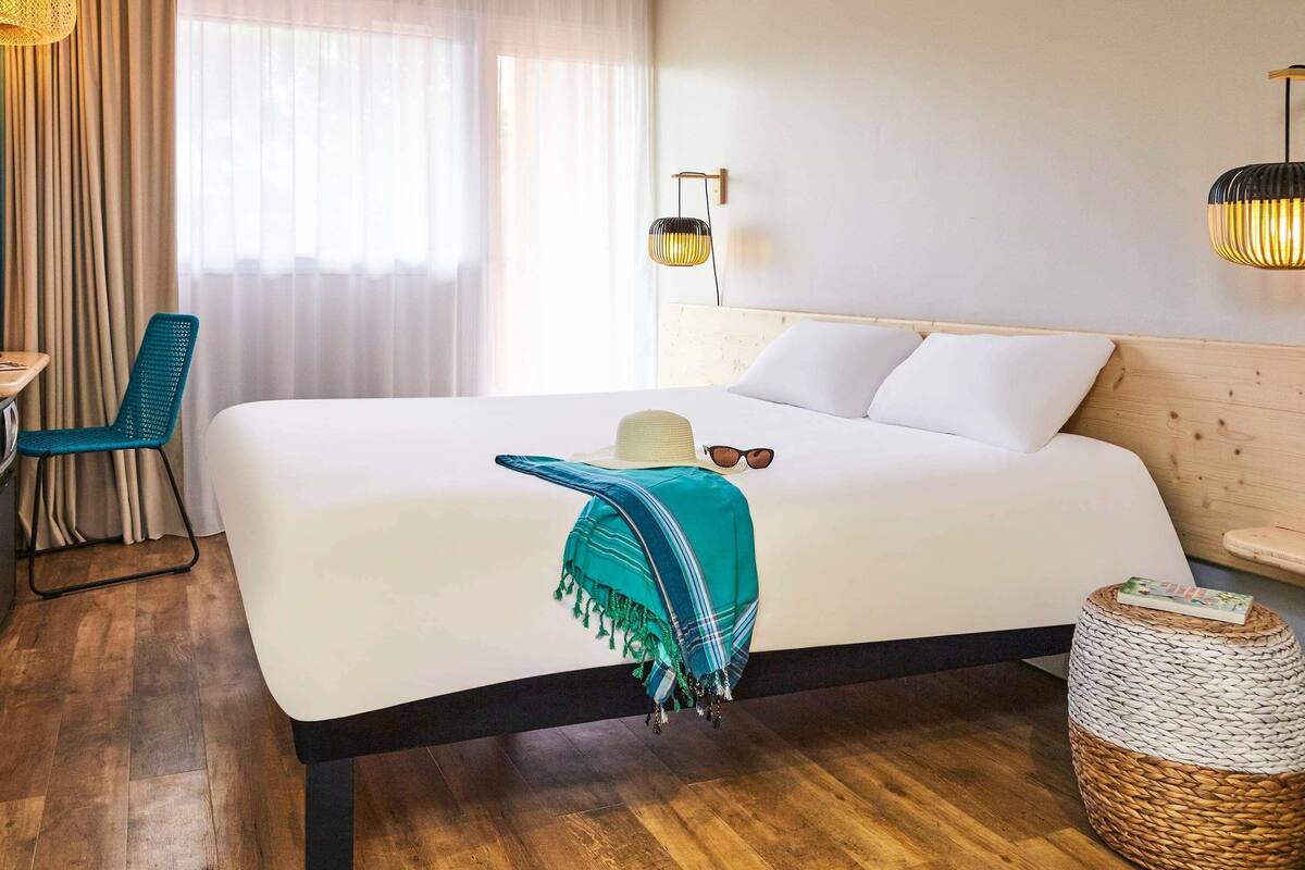 Quarto de hotel com uma cama grande, uma escrivaninha, uma cadeira, um abajur e um banquinho de fibra sintética.