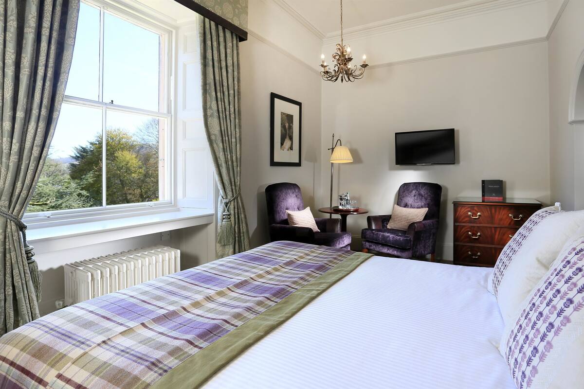 Quarto de hotel com uma cama grande, duas poltronas, uma cômoda, uma televisão e uma janela com cortinas.