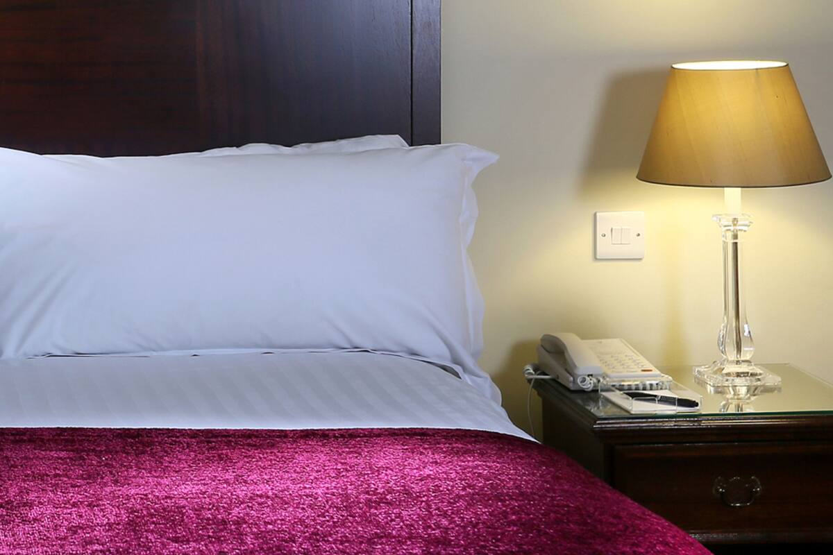 Cama bem arrumada com lençóis brancos, cabeceira de madeira escura, quadro na parede, abajur e telefone na mesa de cabeceira.