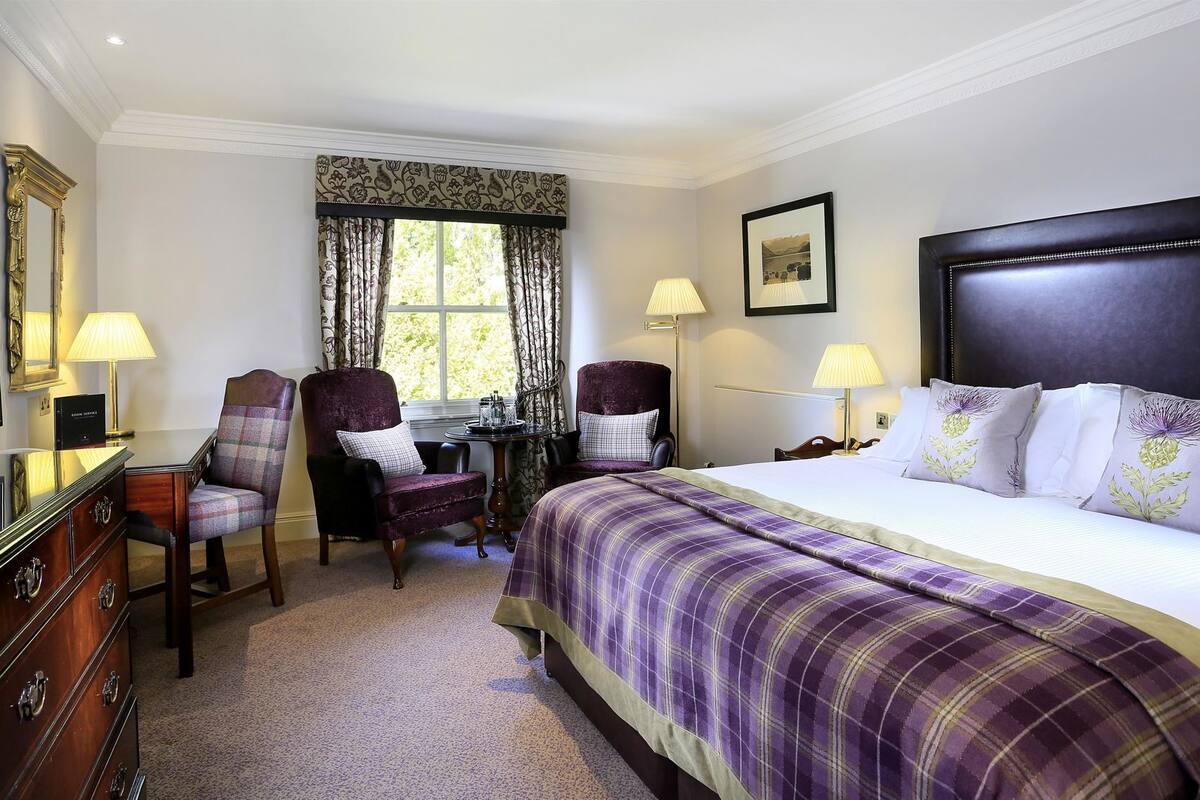 Quarto de hotel com uma cama, duas cadeiras, uma mesa, uma televisão e uma janela com cortinas.