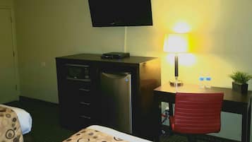 Quarto de hotel com cama, escrivaninha com computador, televisão de tela plana, frigorífico, micro-ondas, candeeiro e planta em vaso.