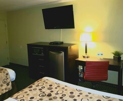 Quarto de hotel com cama, escrivaninha com computador, televisão de tela plana, frigorífico, micro-ondas, candeeiro e planta em vaso.