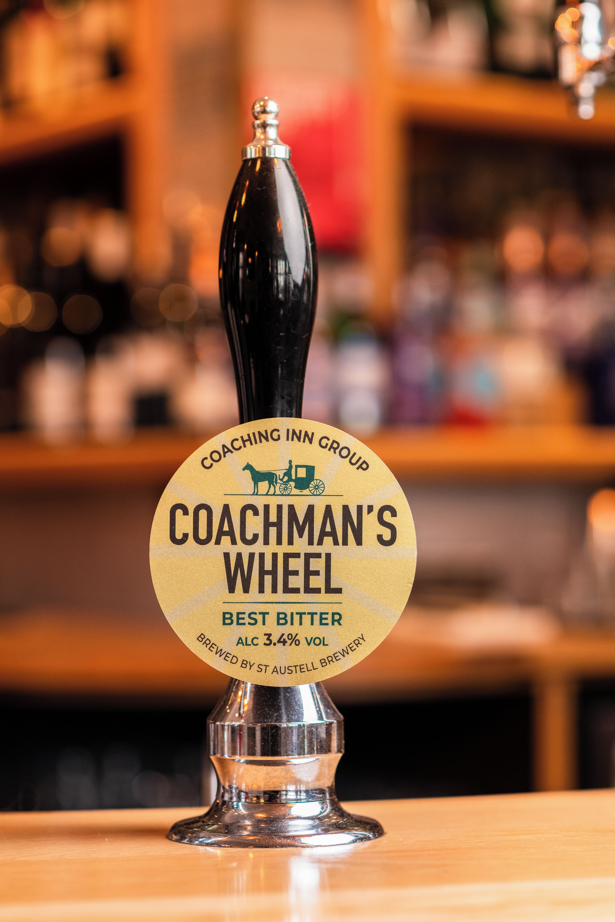 Uma torneira identificada como 'COACHMAN'S WHEEL' com um logotipo e texto indicando que é uma cerveja bitter de qualidade, com 3,4% de teor alcoólico, produzida pela St Austell Brewery.