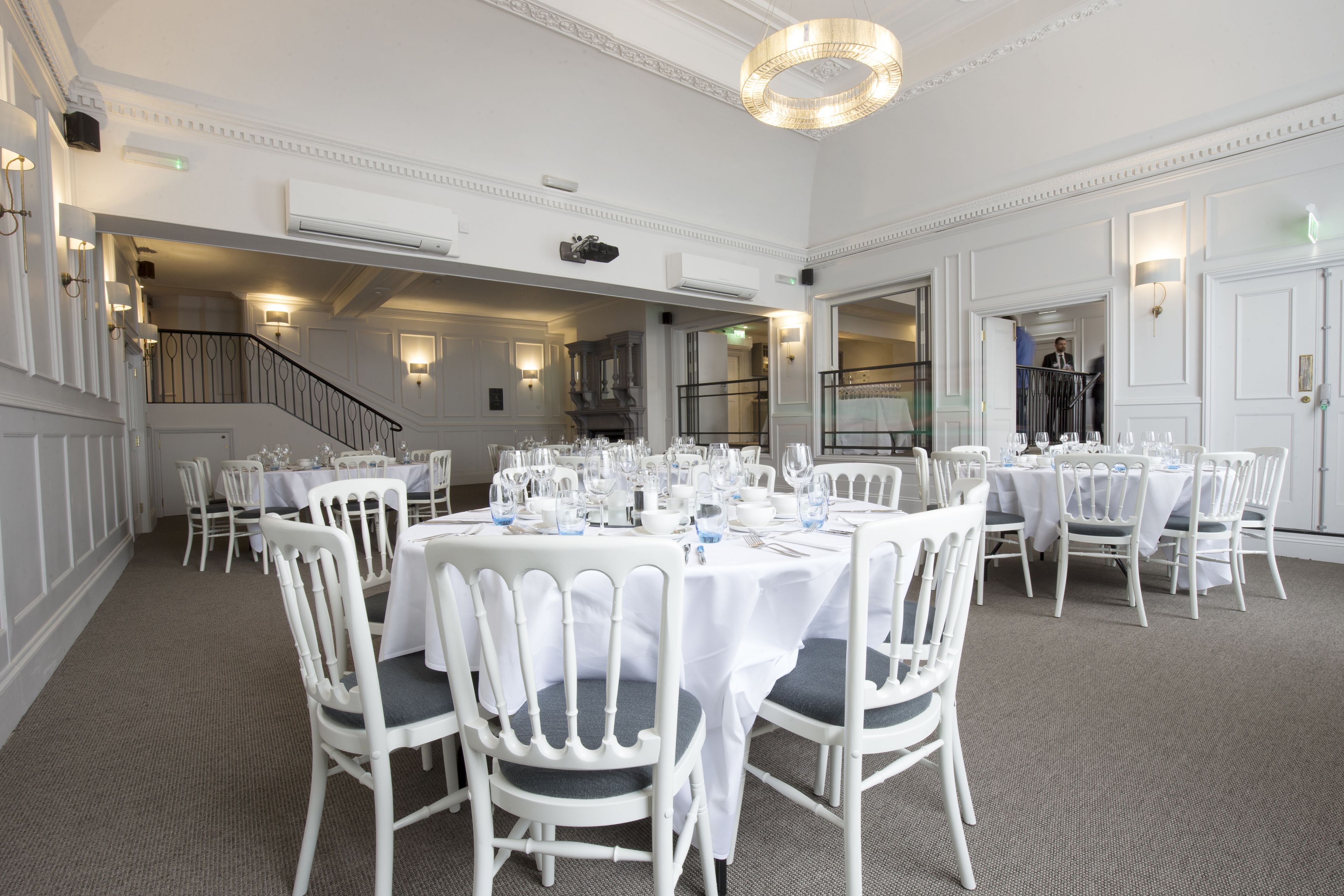Uma sala de jantar formal com mesas e cadeiras brancas, um candelabro e uma escadaria ao fundo.