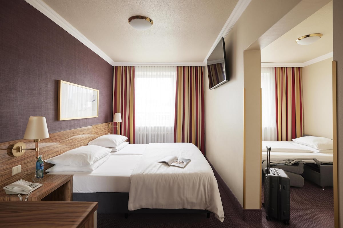 Quarto de hotel com duas camas, uma escrivaninha com luminária, um telefone e uma televisão fixada na parede.