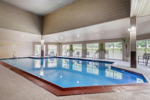 Piscina interior com água cristalina, rodeada por cadeiras e mesas, e grandes janelas que permitem a entrada de luz natural.