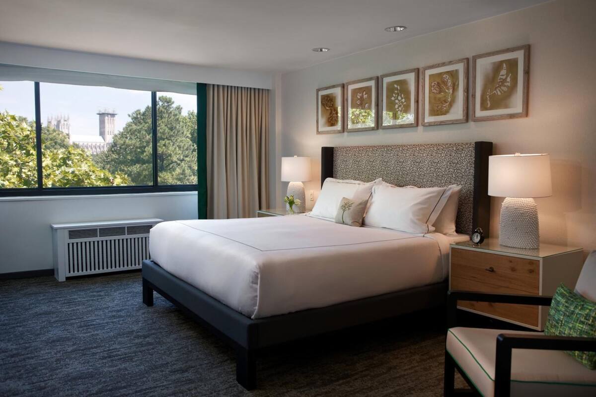 Quarto de hotel com cama grande, luminárias de cabeceira, mesa de cabeceira, uma cadeira e janela com vista para árvores e um edifício.
