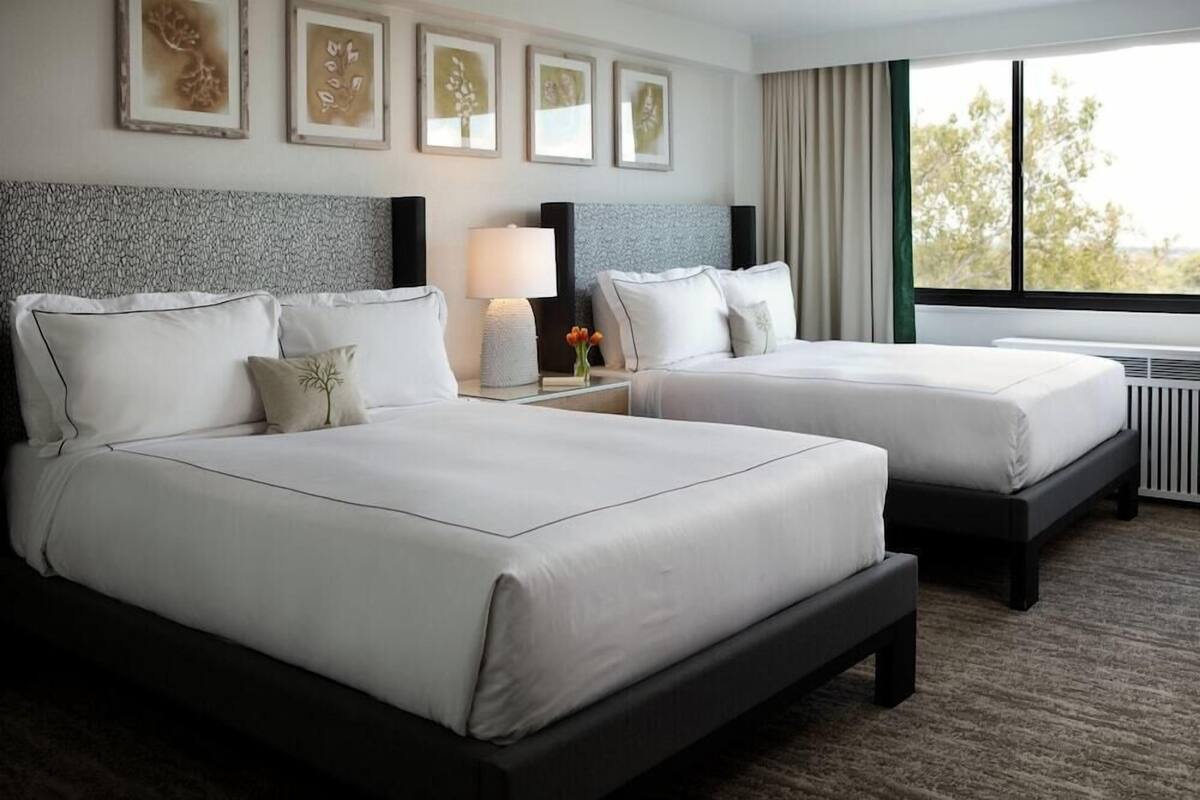 Quarto de hotel com duas camas, uma janela grande e obras de arte na parede.