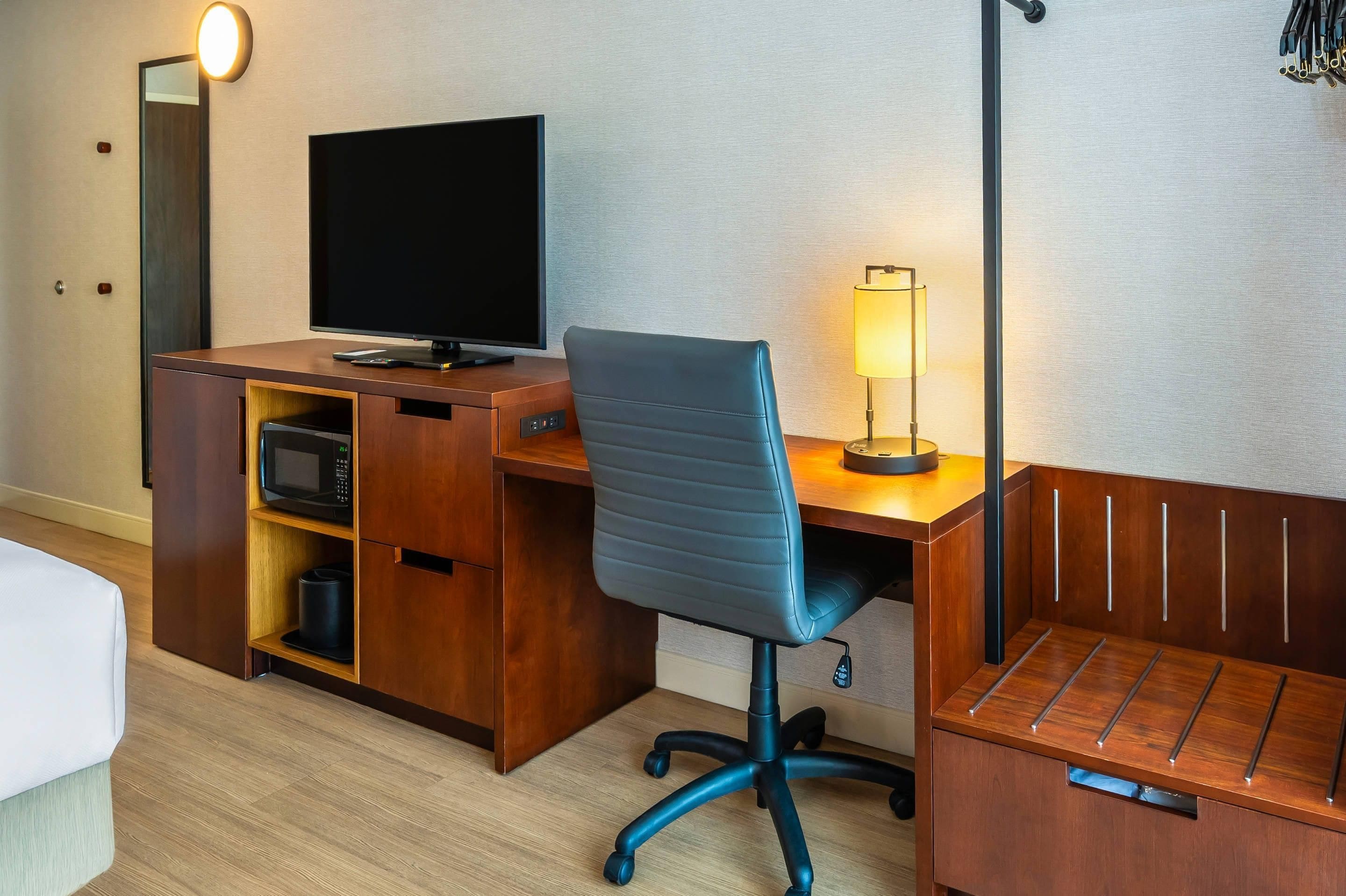 Quarto de hotel com escrivaninha de madeira, televisão, luminária, cadeira e cama.