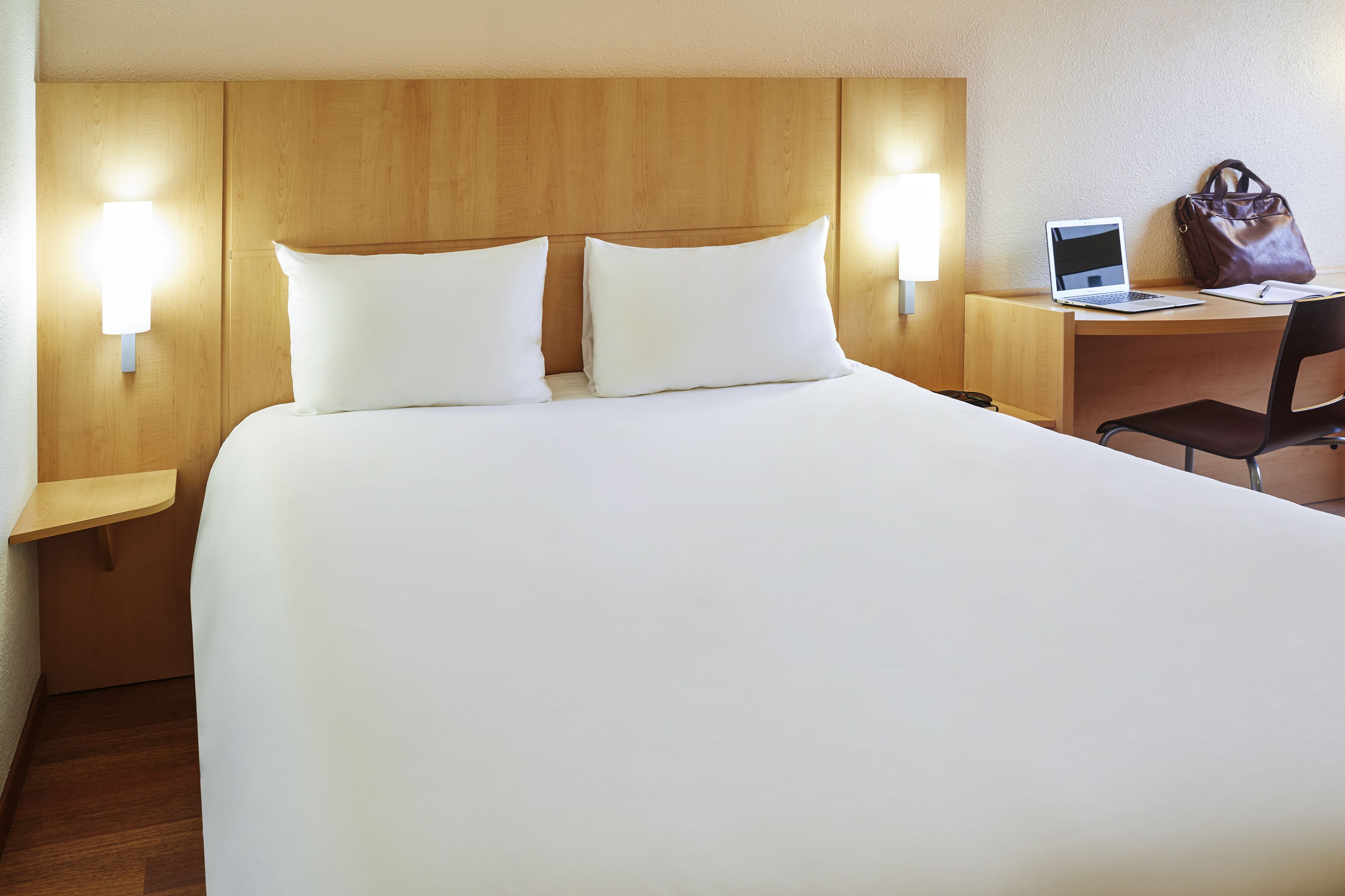 Quarto de hotel com uma cama grande, duas luminárias de cabeceira, uma escrivaninha com um laptop, uma cadeira e uma bolsa.