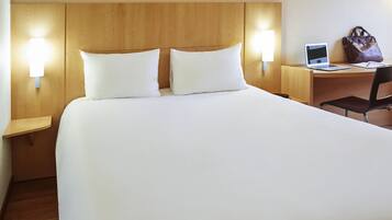 Quarto de hotel com uma cama grande, duas luminárias de cabeceira, uma escrivaninha com um laptop, uma cadeira e uma bolsa.