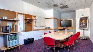 Sala de conferências com uma mesa grande, cadeiras vermelhas, um quadro branco, uma televisão e um frigorífico com bebidas.