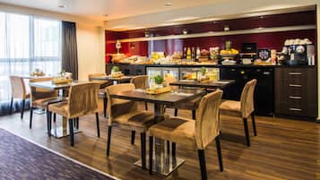 Uma área de jantar de hotel moderna com várias mesas e cadeiras, cozinha com diversos eletrodomésticos e expositores de alimentos, e uma grande janela que proporciona luz natural.