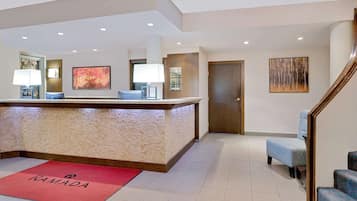 Uma área de recepção com balcão, tapete vermelho com o logotipo "RAMADA", escada e parede com obras de arte emolduradas.