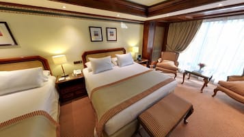 Quarto de hotel com duas camas, área de estar com uma cadeira e uma mesa, e uma janela grande com cortinas.