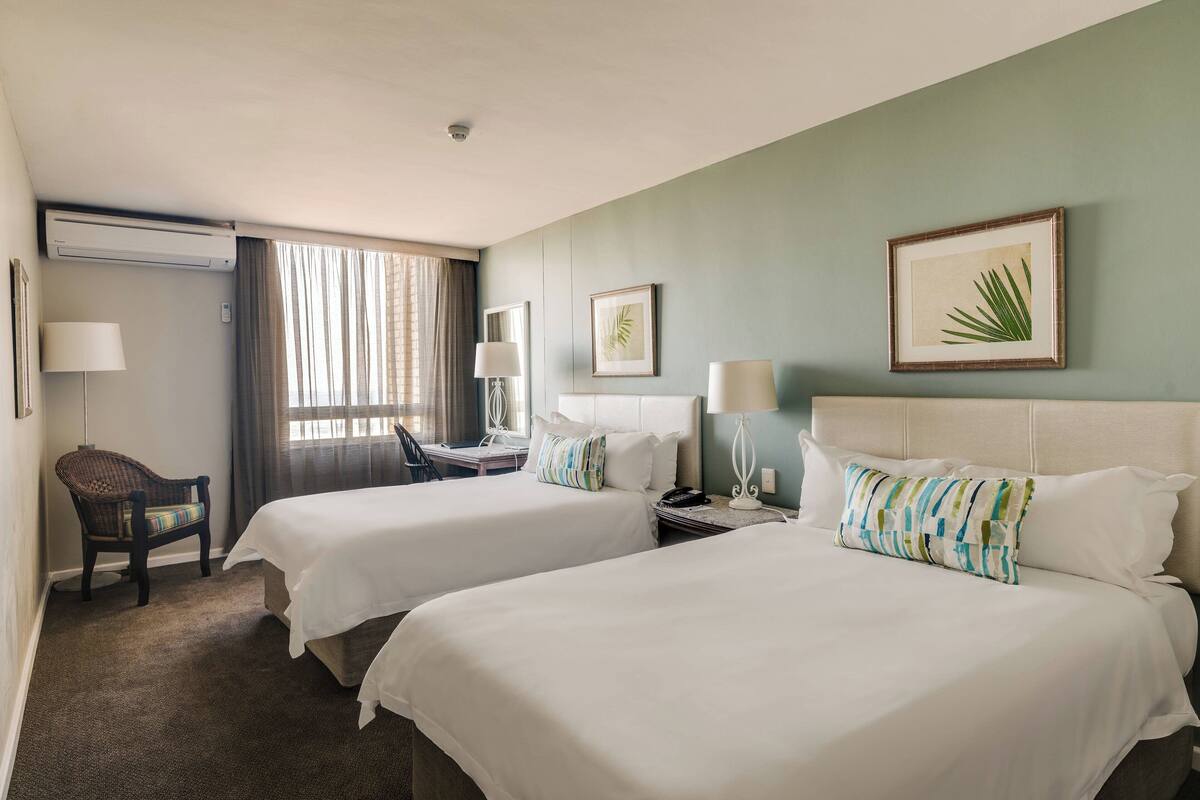 Quarto de hotel com duas camas, uma mesa, uma cadeira e uma janela com cortinas.