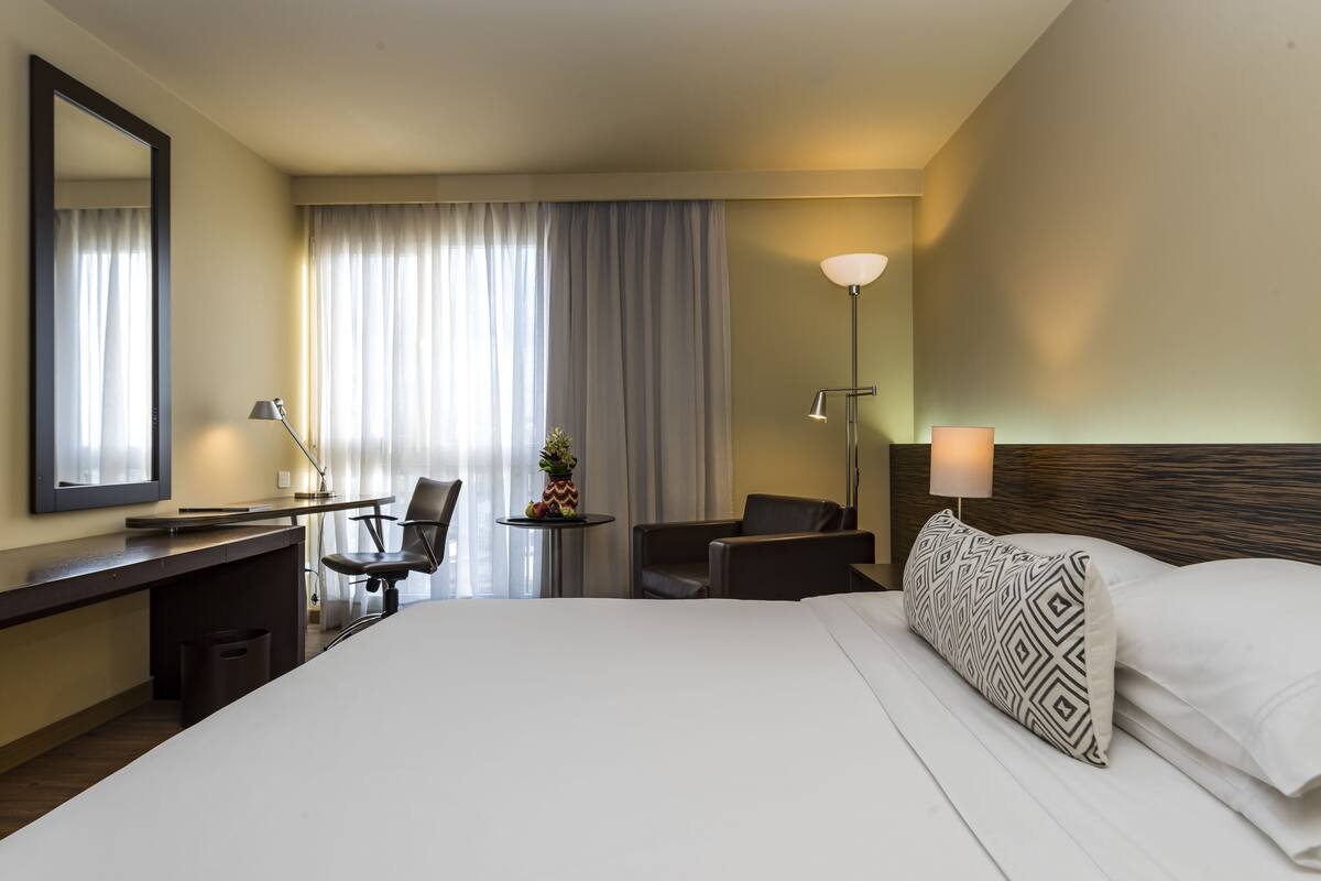 Quarto de hotel com uma cama grande, uma escrivaninha, duas cadeiras, uma mesinha de centro com uma planta e uma janela ampla com cortinas.