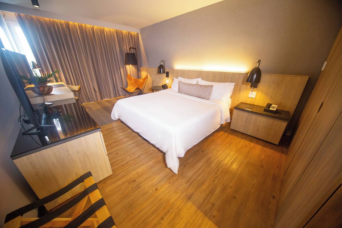 Quarto de hotel com uma cama grande, uma escrivaninha com um computador, uma cadeira e uma mesa de cabeceira com abajur.