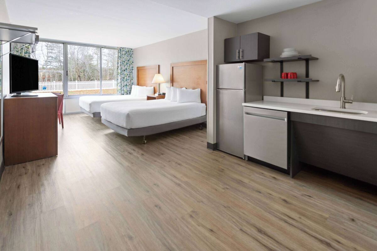 Quarto de hotel com duas camas, televisão, escrivaninha, frigorífico e cozinha equipada com pia e prateleiras.