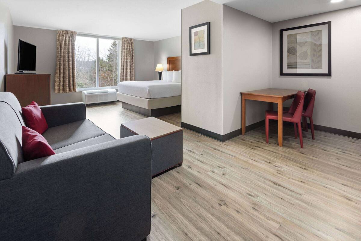 Quarto de hotel moderno com um sofá cinzento, uma mesa de jantar de madeira com duas cadeiras, uma cama com roupa de cama branca, uma TV de tela plana sobre um móvel de madeira e uma janela grande com cortinas.