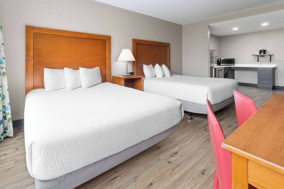Quarto de hotel com duas camas, uma escrivaninha, uma cadeira e uma cozinha compacta.