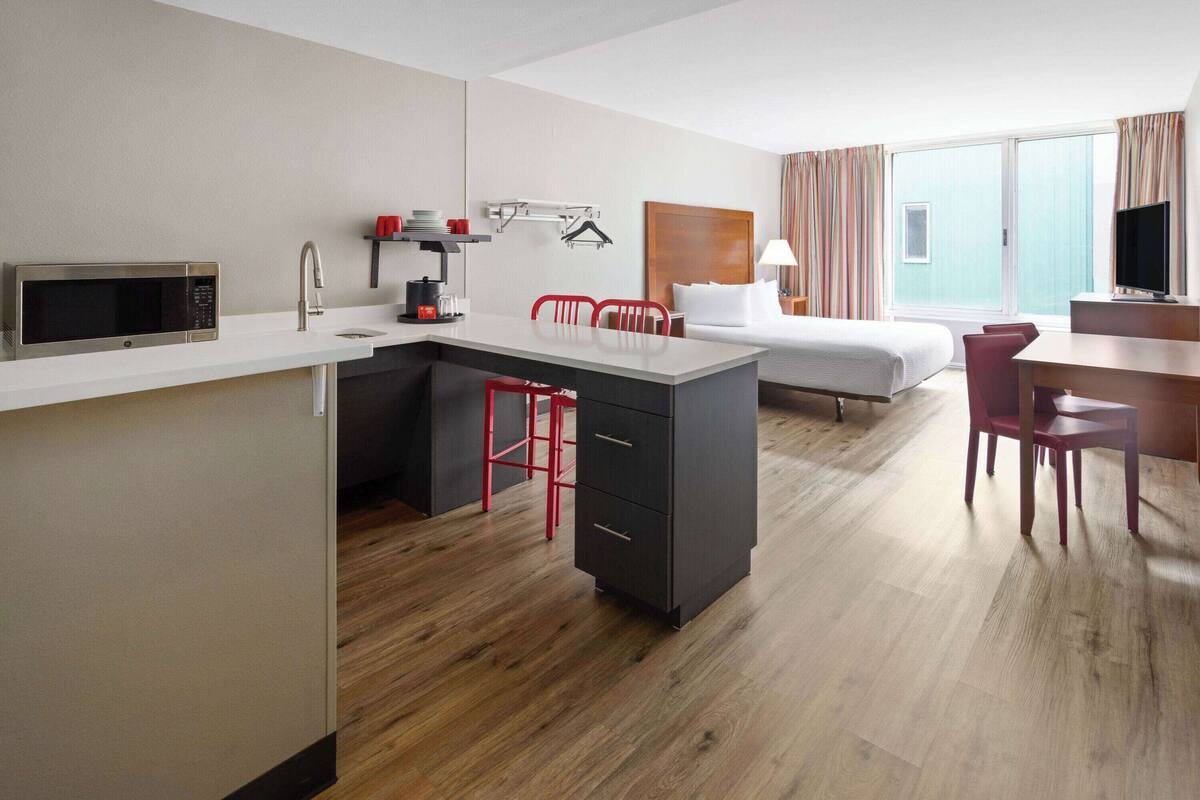 Quarto de hotel moderno com cozinha compacta, cama, escrivaninha e televisão.